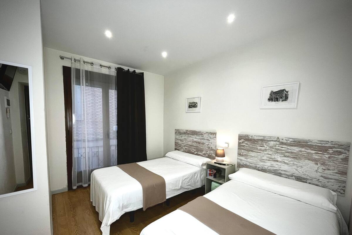 Quarto de hotel com duas camas, mesa de cabeceira com abajur, janela com cortinas e um espelho.
