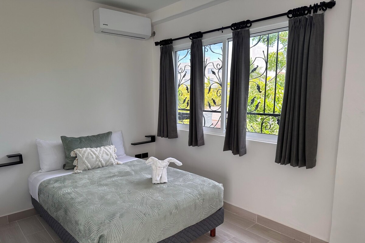 Cama bem arrumada com colcha verde estampada, um travesseiro branco e uma toalha branca ao lado. Há uma janela com cortinas escuras e vista para o verde lá fora.