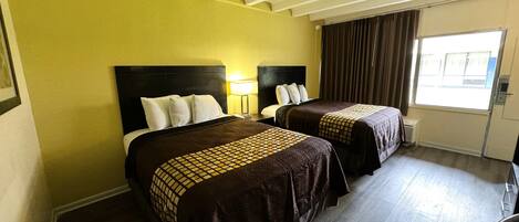 Quarto de hotel com duas camas, uma mesa de cabeceira, uma luminária e uma janela com cortinas.
