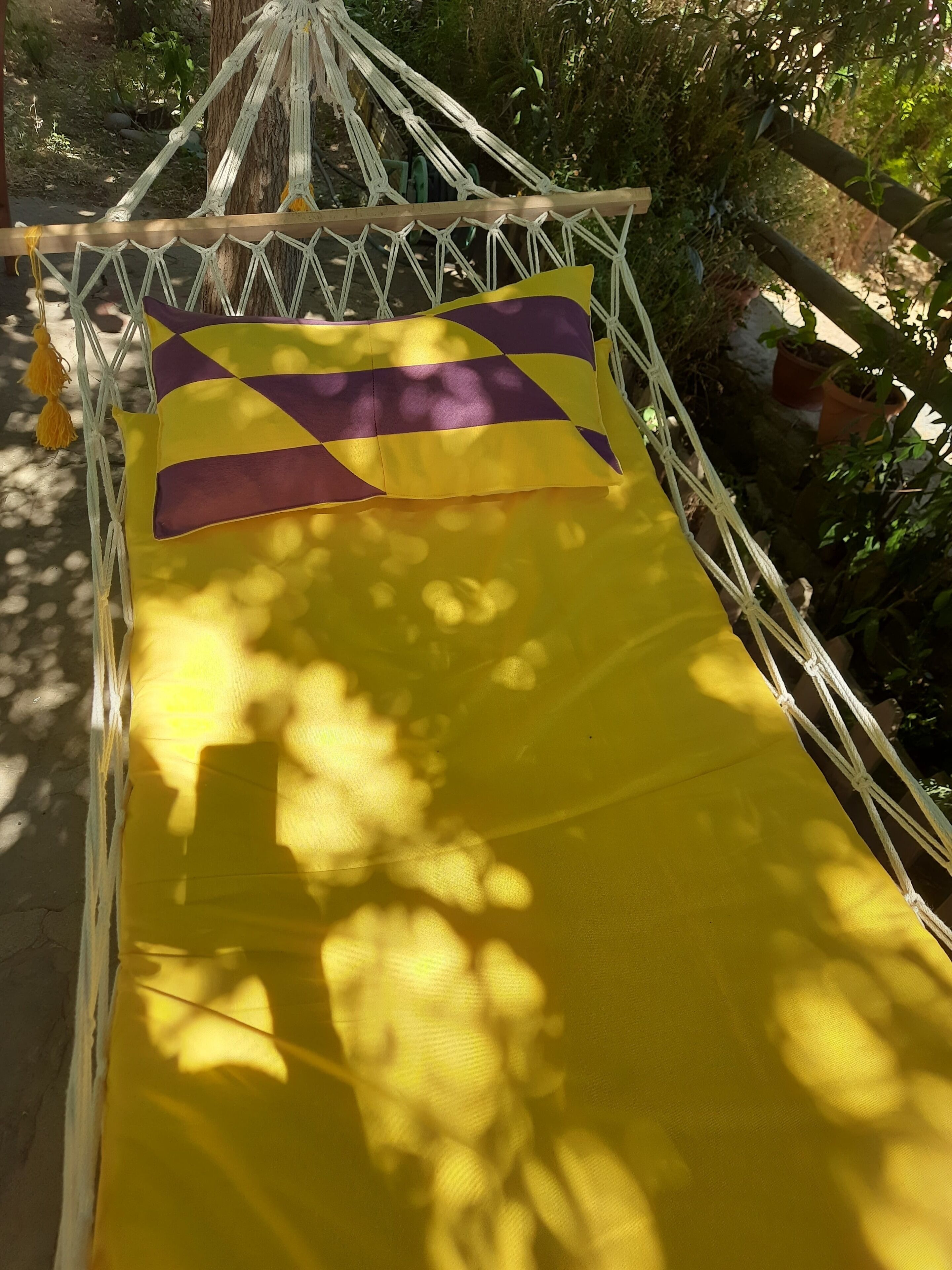 Uma rede com um travesseiro listrado em amarelo e roxo, cercada por árvores e folhagem.