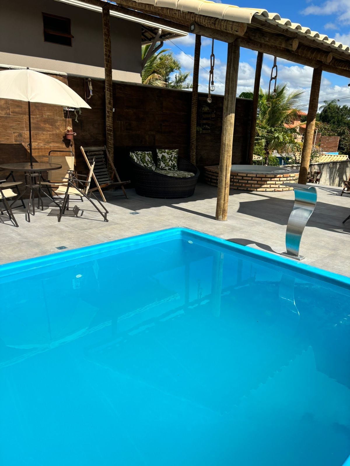 Piscina com pergolado, área de jantar com cadeiras e mesa, e espreguiçadeira com almofadas.