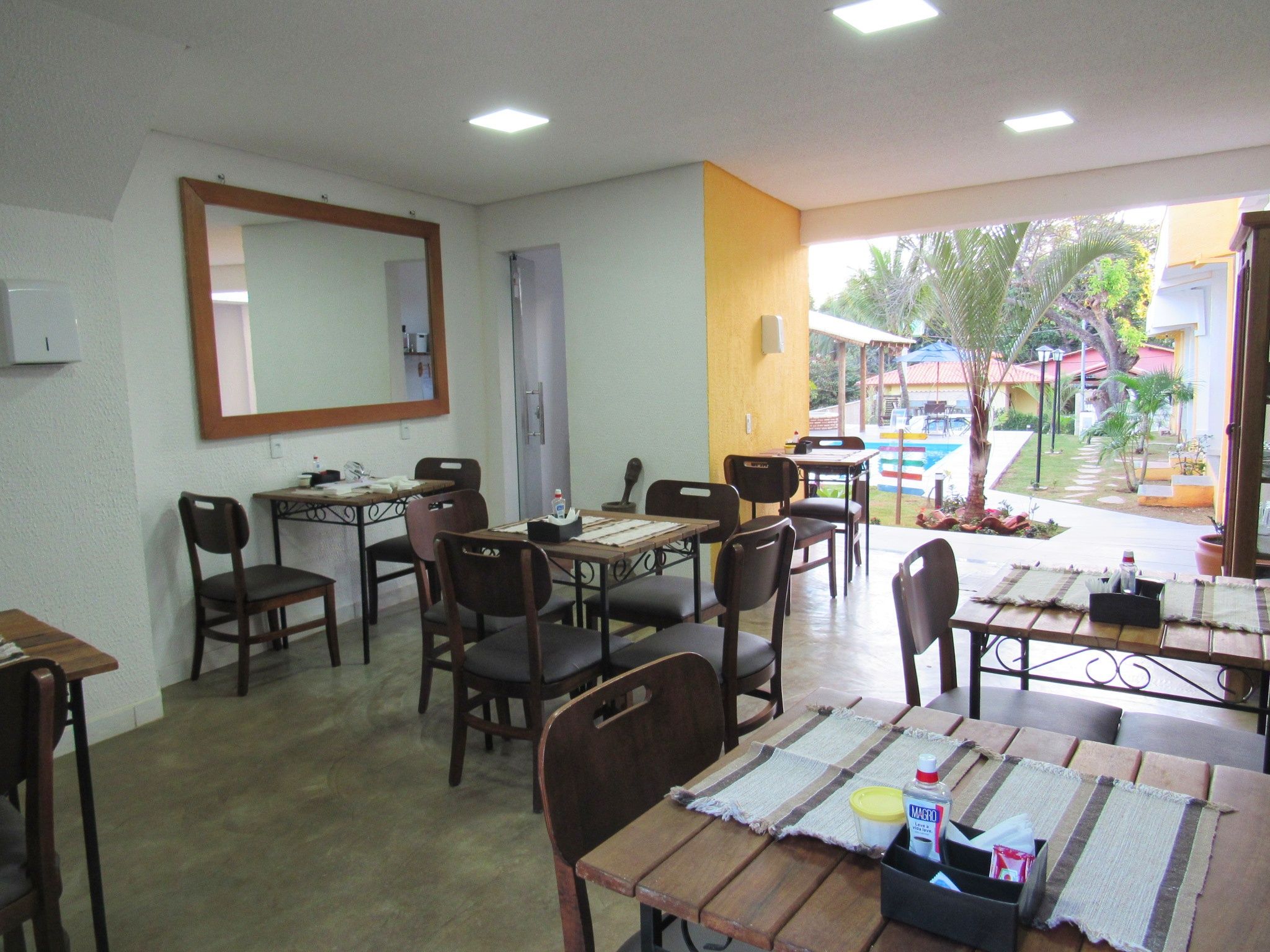Espaço de refeições com várias mesas e cadeiras, um espelho grande na parede e vista para a área da piscina.