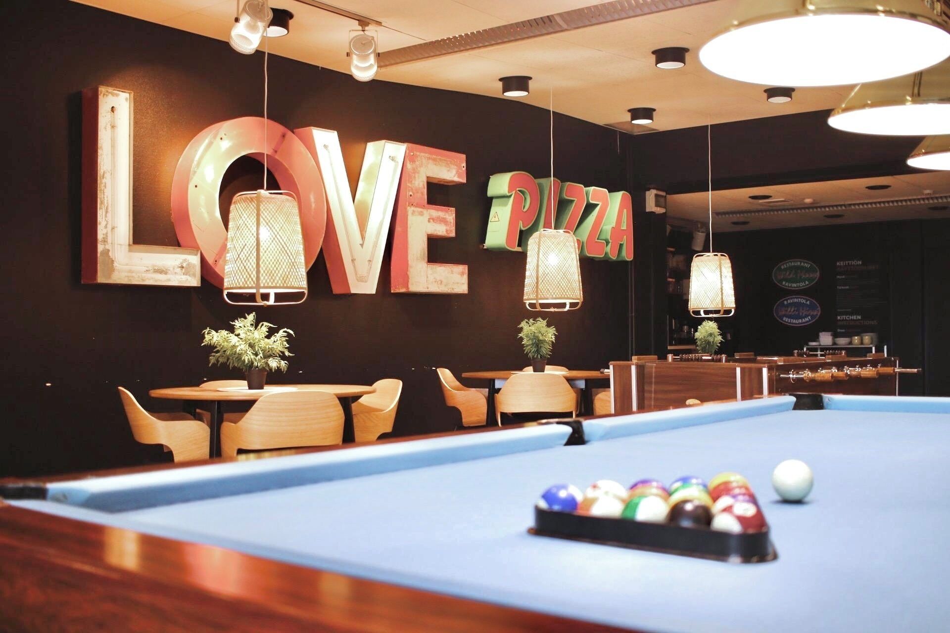 Uma mesa de bilhar com bolas de bilhar, uma mesa de sinuca e uma parede com letras iluminadas que formam a frase 'LOVE PIZZA'.