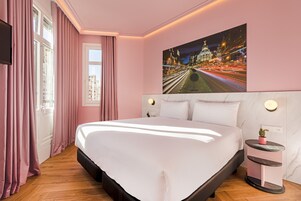 Quarto moderno com cama grande, cortinas rosa, pintura de paisagem urbana e vista noturna de uma rua iluminada.
