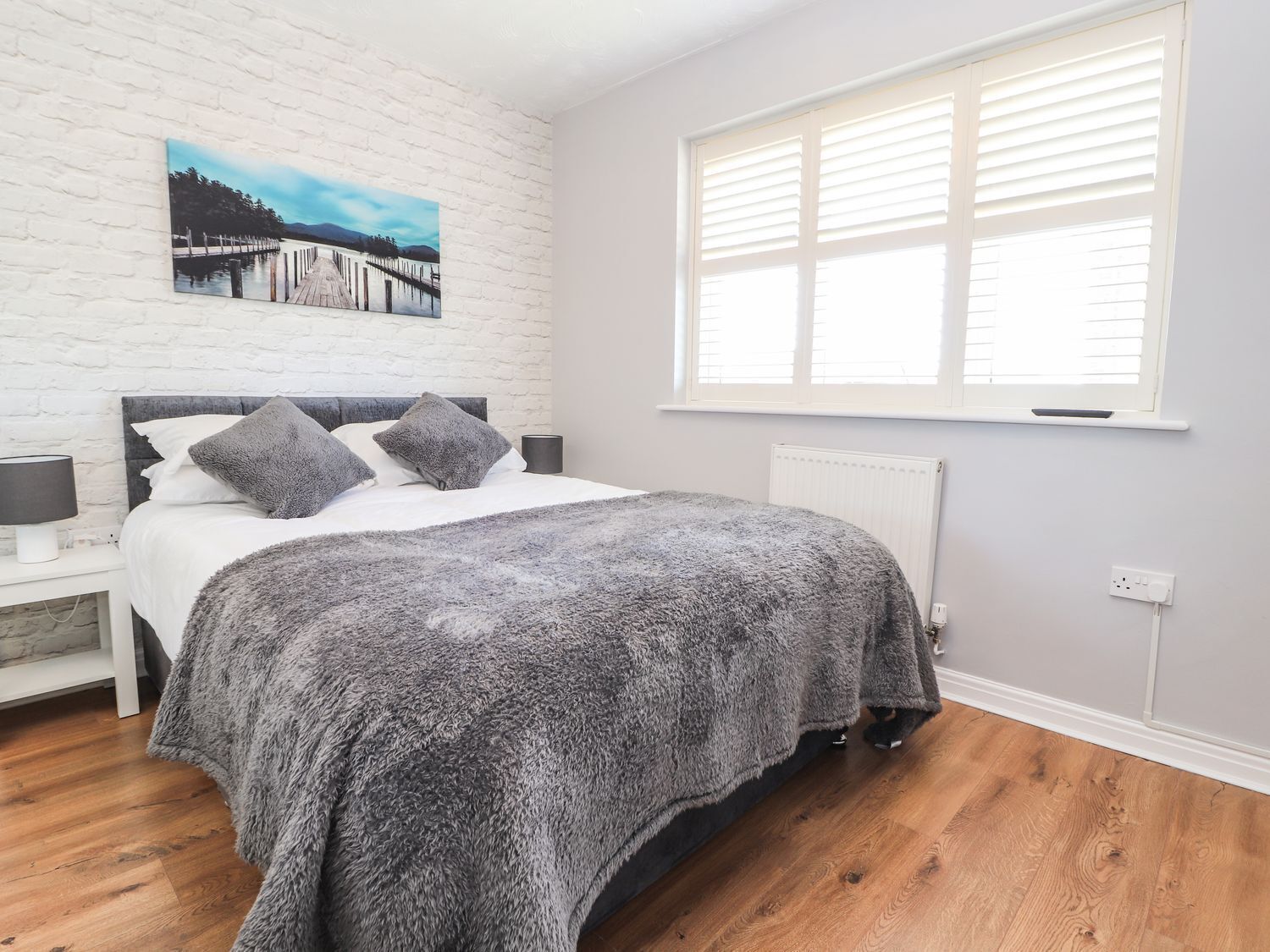 Quarto com parede de tijolos brancos, cama com roupa de cama cinza, janela com persianas e um quadro na parede.