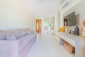 Uma sala de estar moderna com um sofá, uma TV na parede e uma área de cozinha ao fundo.