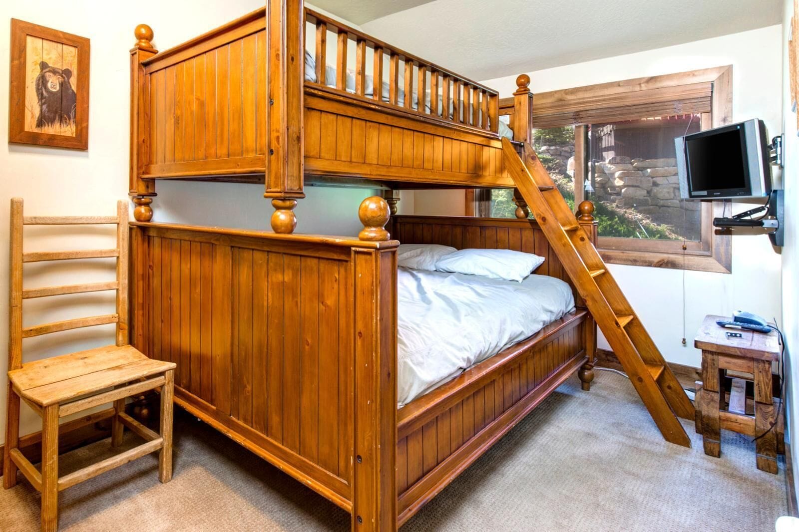 Uma cama beliche com escada de madeira, uma cadeira de madeira, uma televisão em um suporte e uma janela com vista para uma parede de pedra e vegetação.