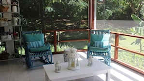 Quarto com duas cadeiras azuis de vime, uma mesa branca e uma janela grande com vista para um jardim.