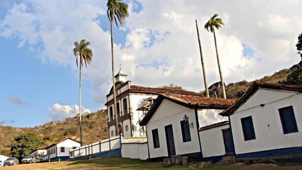 Fileira de edifícios brancos com uma torre de sino, cercada por palmeiras e uma paisagem montanhosa.