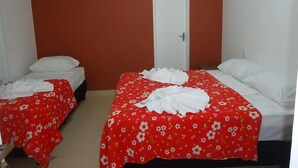 Quarto com duas camas, cada uma com colcha floral vermelha e almofadas brancas. Uma porta branca está centralizada na parede do fundo.