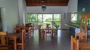 Uma sala de jantar ampla com mesas e cadeiras de madeira, grandes janelas e vista para o verde lá fora.