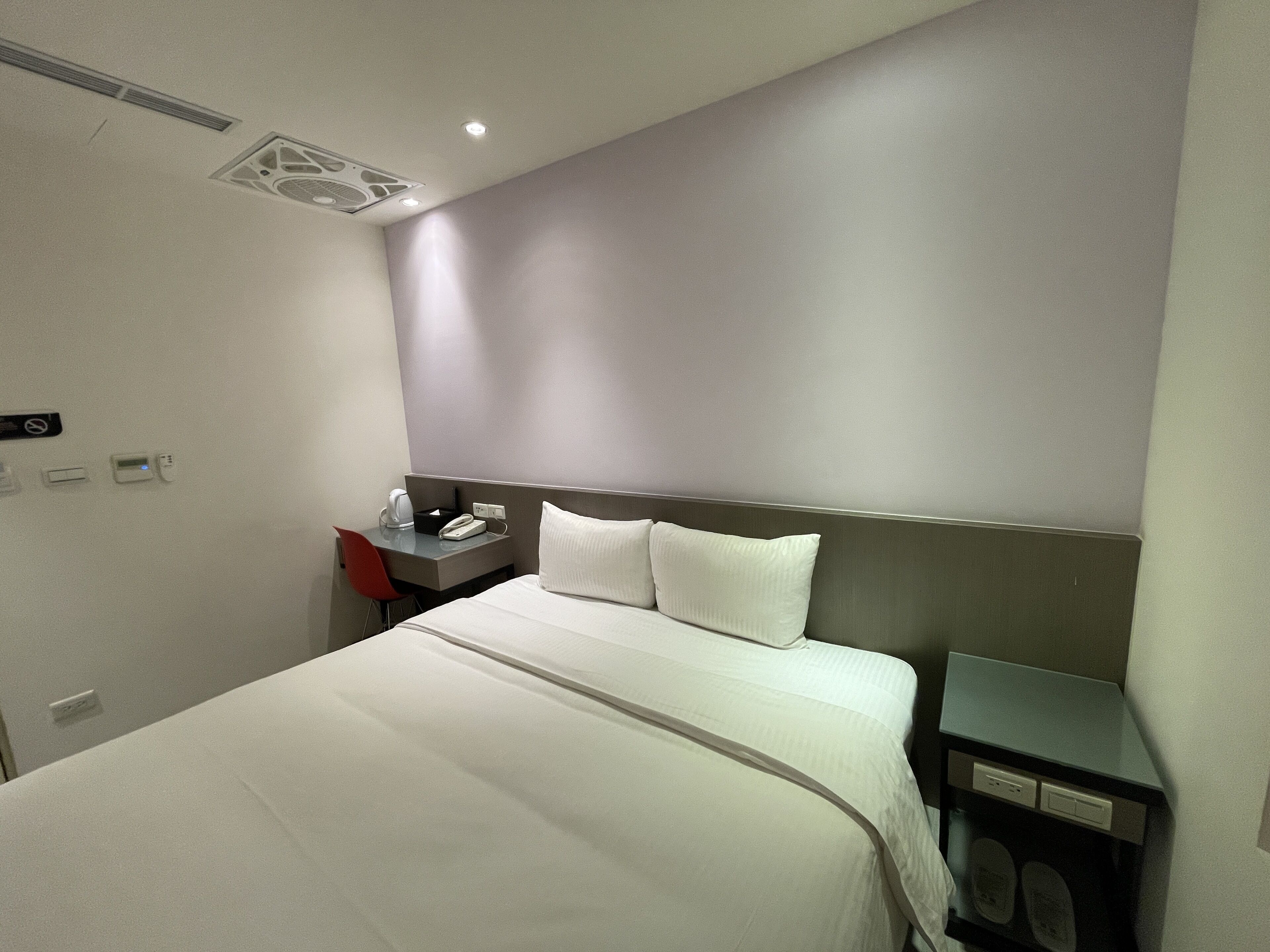 Quarto de hotel com cama, mesa de cabeceira, uma cadeira vermelha e telefone fixado na parede.