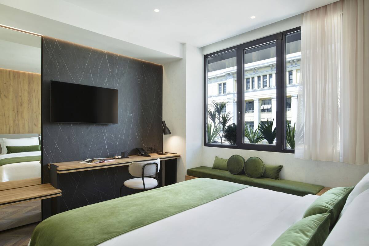 Quarto de hotel moderno com uma cama grande, uma escrivaninha com cadeira, televisão de tela plana, janela com vista para edifícios e um sofá verde com almofadas.