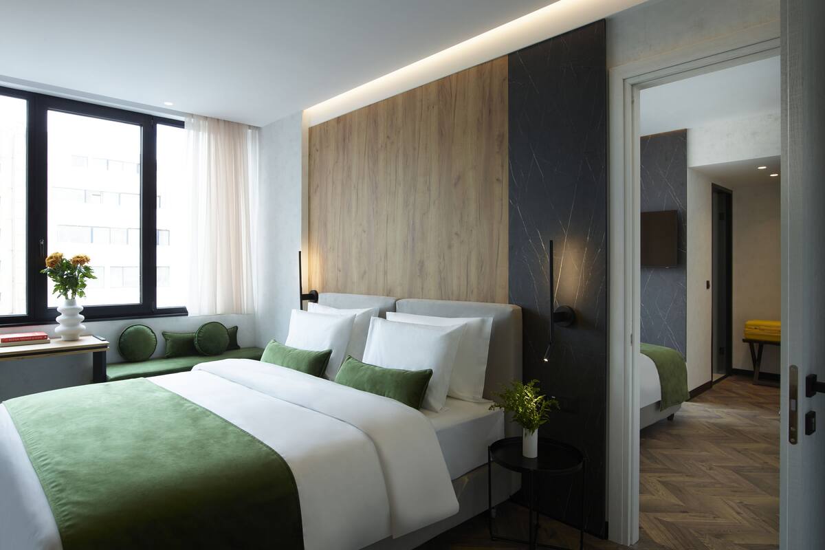 Quarto de hotel moderno com uma cama grande, uma mesa de cabeceira com um vaso e vista da cidade por grandes janelas.