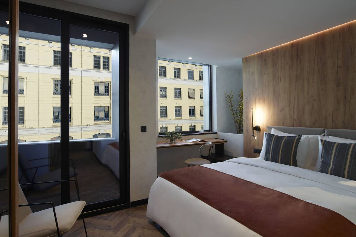 Quarto de hotel moderno com uma cama grande, uma escrivaninha e vista da cidade através de janelas do chão ao teto.