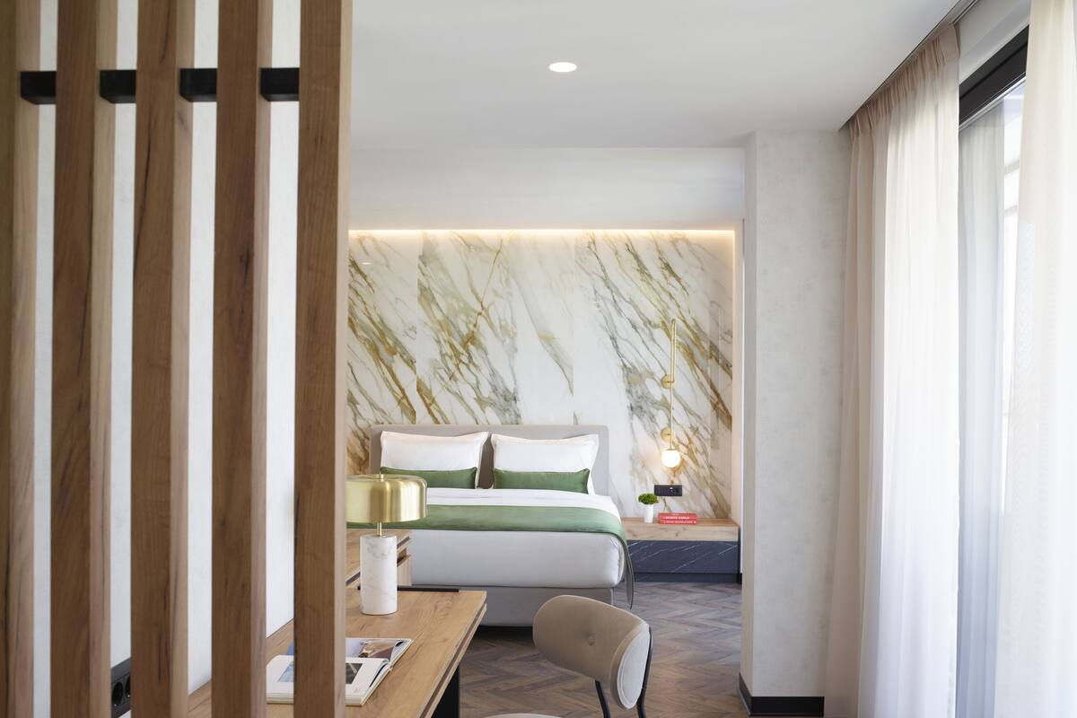 Quarto de hotel moderno com parede em padrão marmorizado, cama bem arrumada com roupa de cama verde, escrivaninha de madeira com cadeira e mesa de cabeceira com abajur.