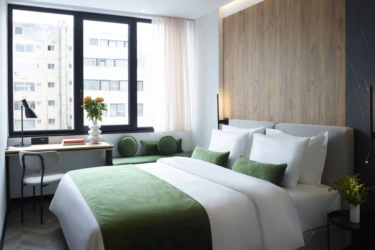 Quarto de hotel moderno com uma cama grande, uma escrivaninha com cadeira, uma janela com cortinas e vista para edifícios.