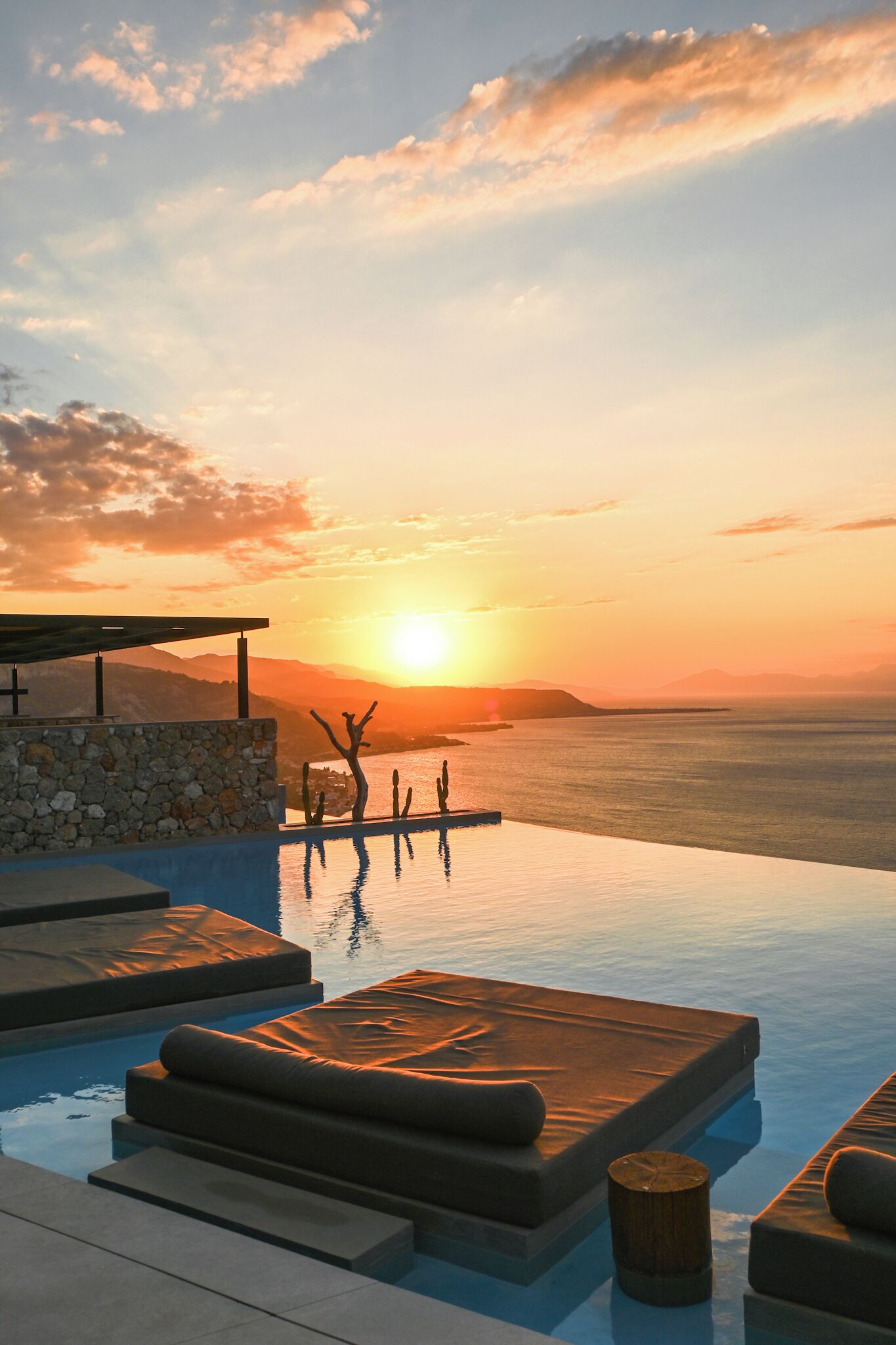 Uma piscina infinita com espreguiçadeiras e uma parede de pedra, com vista para o pôr do sol sobre a água.