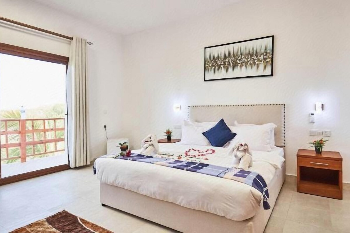 Quarto com cama grande, mesa de cabeceira, luminária na parede e janela com vista.