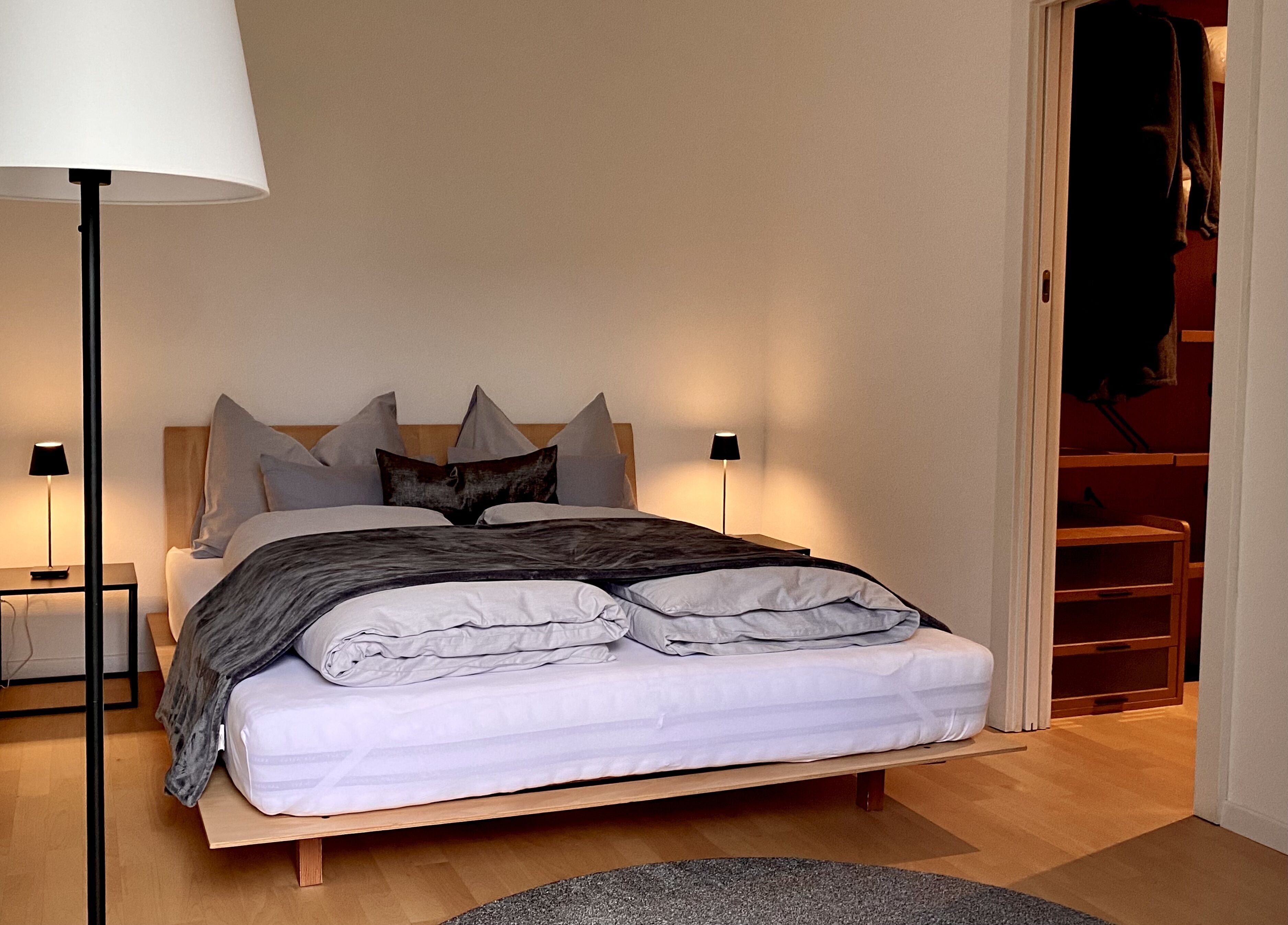 Cama bem arrumada com edredom cinza e branco, abajur e cabeceira de madeira.
