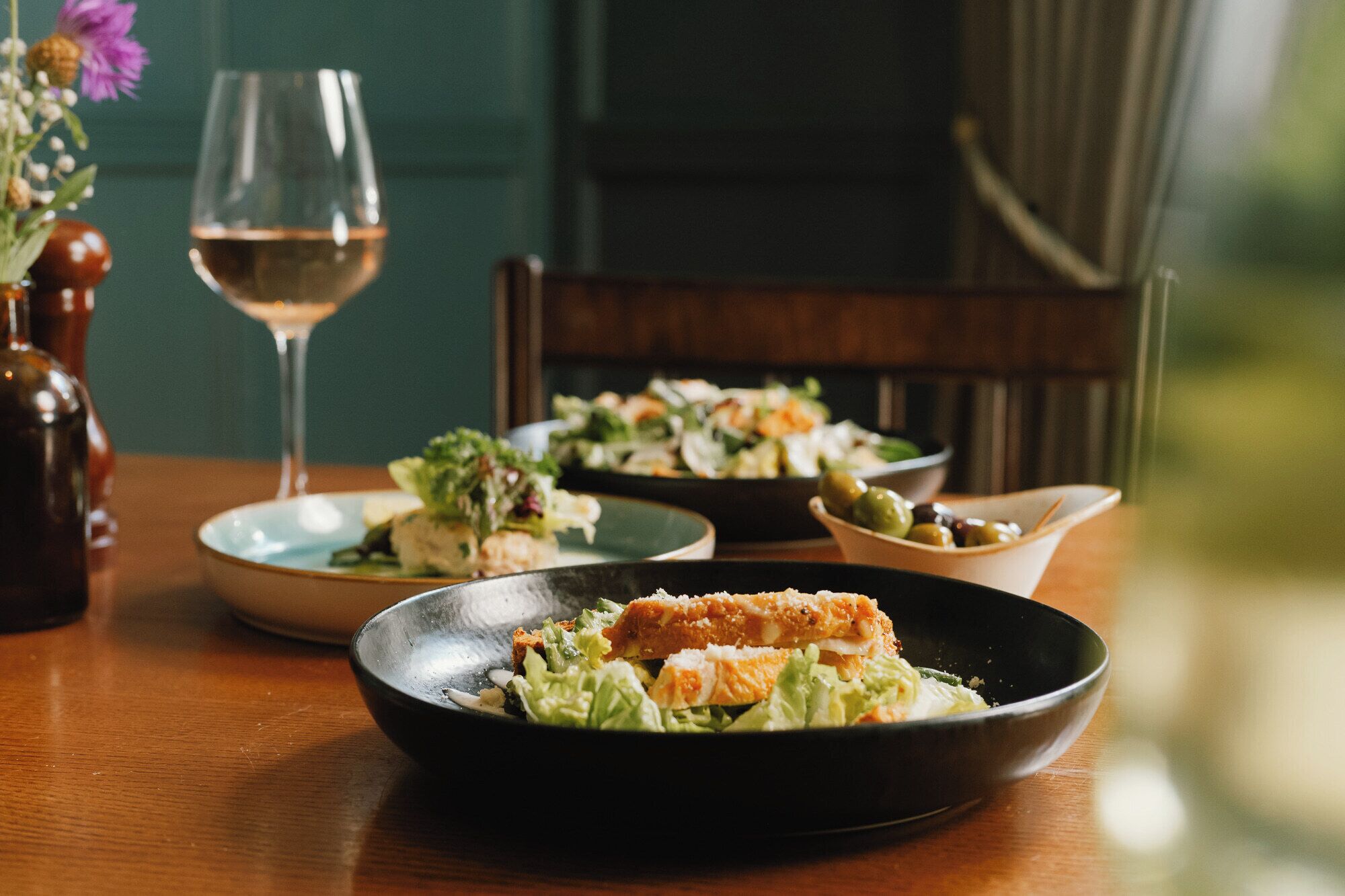 Uma mesa de jantar com uma salada, um copo de vinho e um vaso com flores.