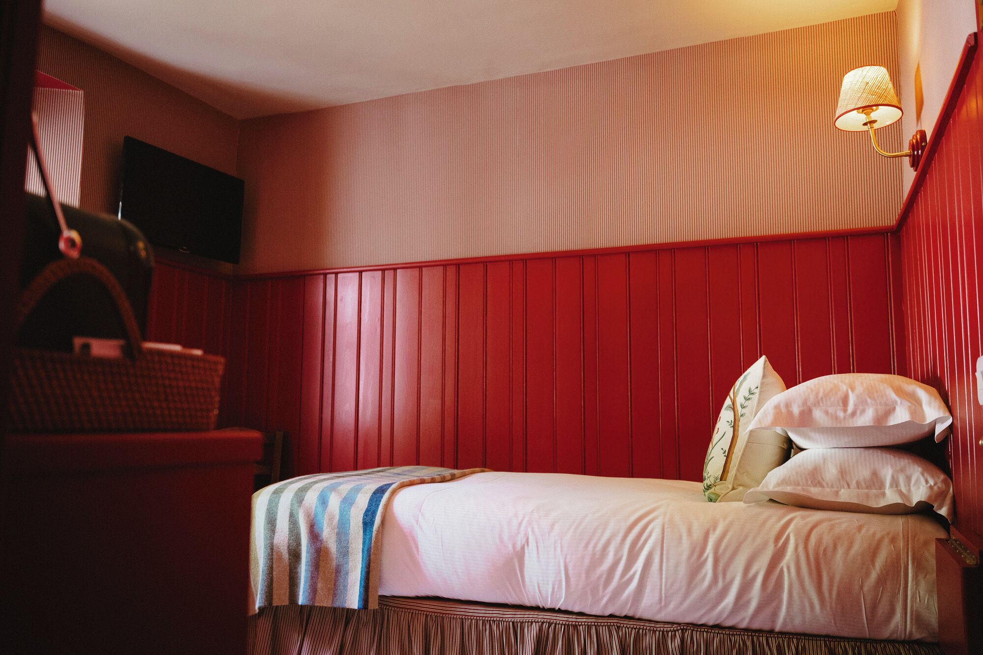 Quarto com cama, almofadas, luminária, televisão e parede vermelha.