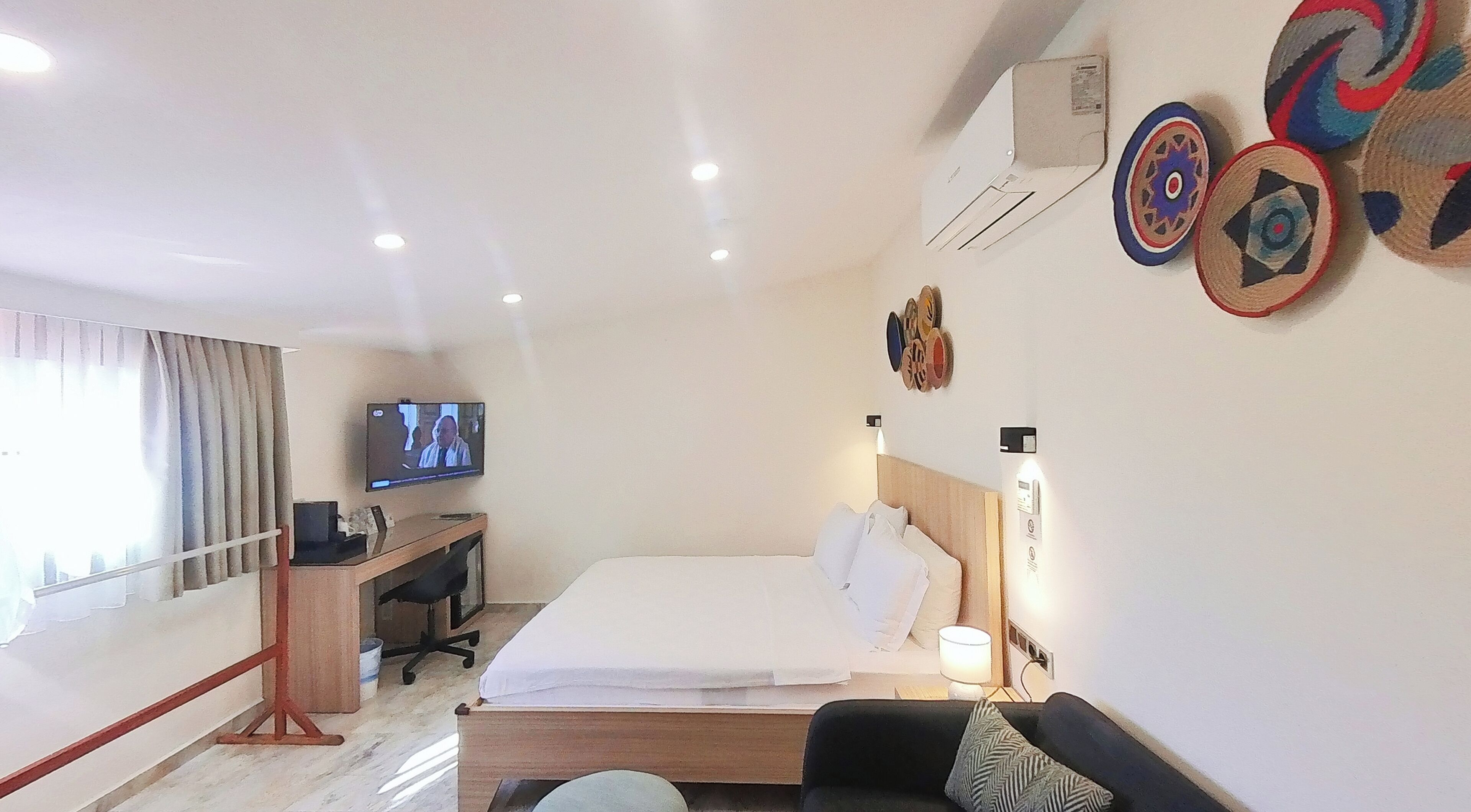Quarto de hotel com cama, mesa de cabeceira, luminária, TV, escrivaninha, cadeira e arte decorativa na parede.