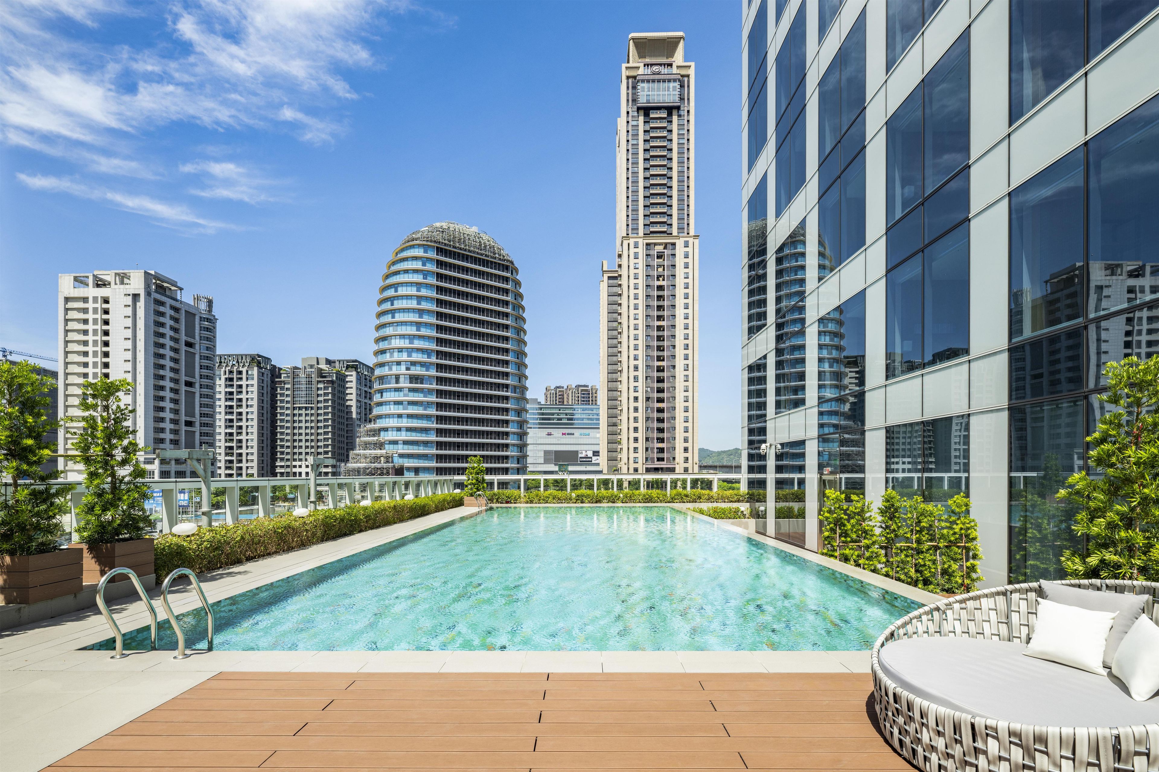 Piscina no terraço com deck de madeira, rodeada por edifícios modernos e vegetação.