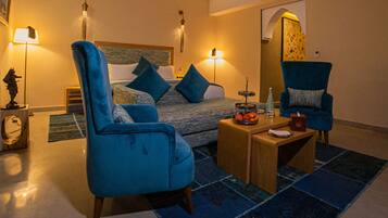 Quarto de hotel com uma cama, duas poltronas azuis, uma pequena mesa lateral de madeira e um espelho.