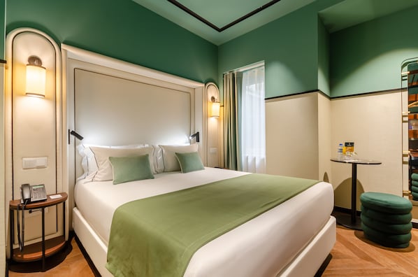 Quarto de hotel com uma cama grande, abajures, uma pequena mesa com uma garrafa e um banquinho verde.