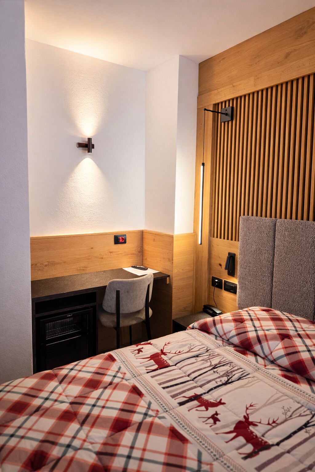 Quarto de hotel com cama, escrivaninha, cadeira, luminária na parede, telefone e painel de parede em madeira.