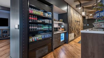Máquina de bebidas moderna abastecida com diversas opções, um pequeno frigorífico e um balcão com petiscos.
