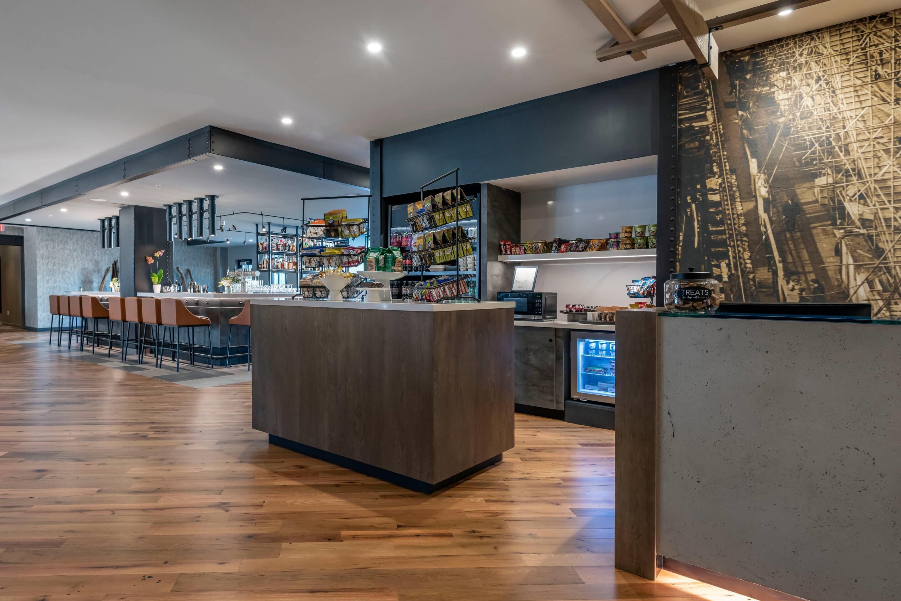 Uma área de bar moderna com um balcão central de madeira, assentos e prateleiras repletas de lanches e bebidas.