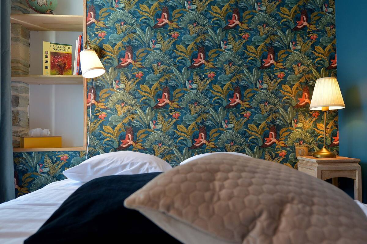 Quarto com papel de parede floral, cama com roupa de cama branca e bege, e duas luminárias de cabeceira.