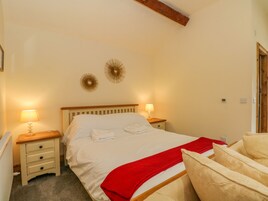Uma cama bem arrumada com lençóis brancos e um cobertor vermelho, duas luminárias de cabeceira, uma mesinha de cabeceira de madeira e um objeto decorativo fixado na parede.