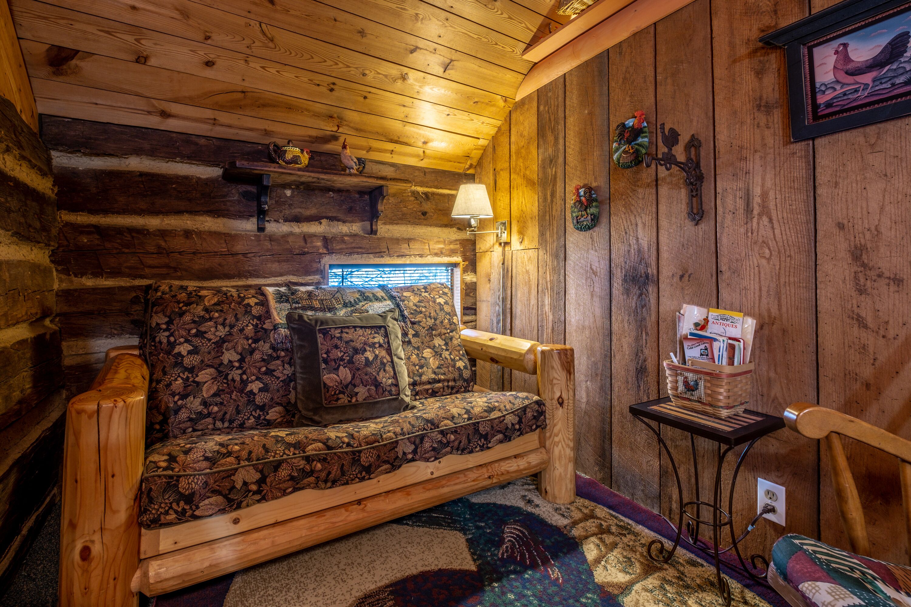 Interior acolhedor de uma cabana com parede de toras de madeira, um sofá estampado com flores, uma mesa pequena com abajur e um quadro na parede.