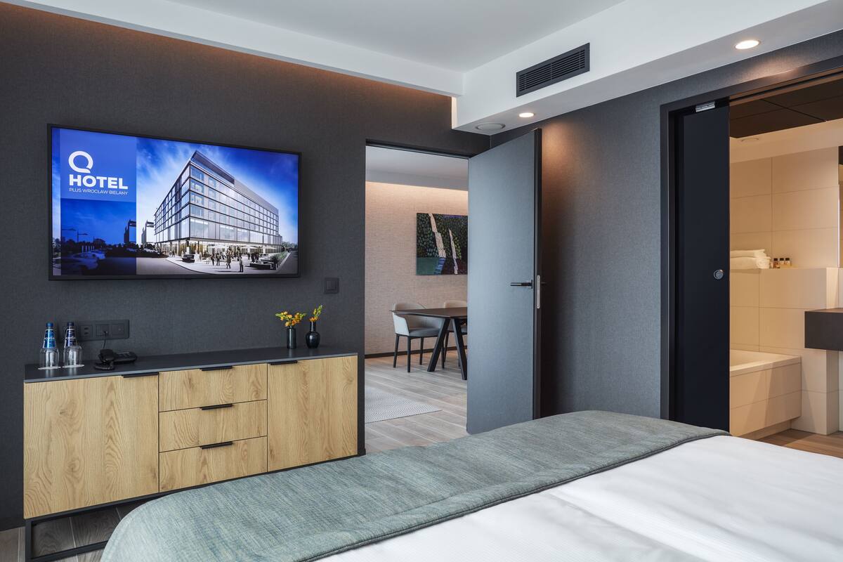 Quarto de hotel moderno com uma cama grande, televisão montada na parede, cômoda de madeira e porta que dá para um banheiro.