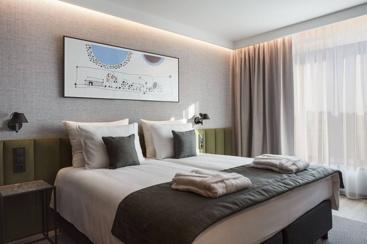 Quarto de hotel moderno com uma cama grande, cabeceira com painel verde e uma obra de arte abstrata emoldurada na parede, além de uma janela com cortinas finas.