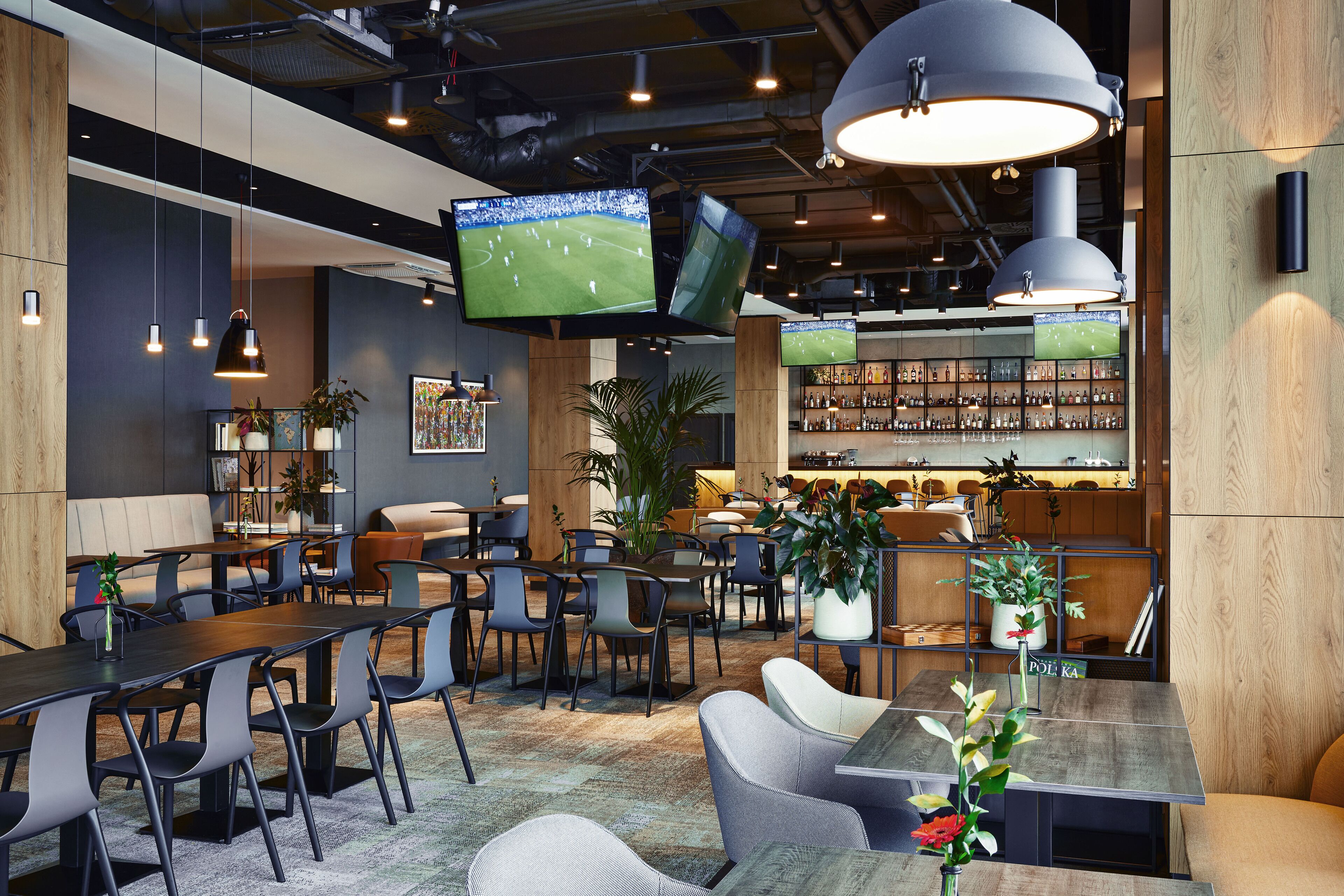Um restaurante moderno com várias mesas e cadeiras, luminárias pendentes grandes e televisões de tela plana exibindo esportes.