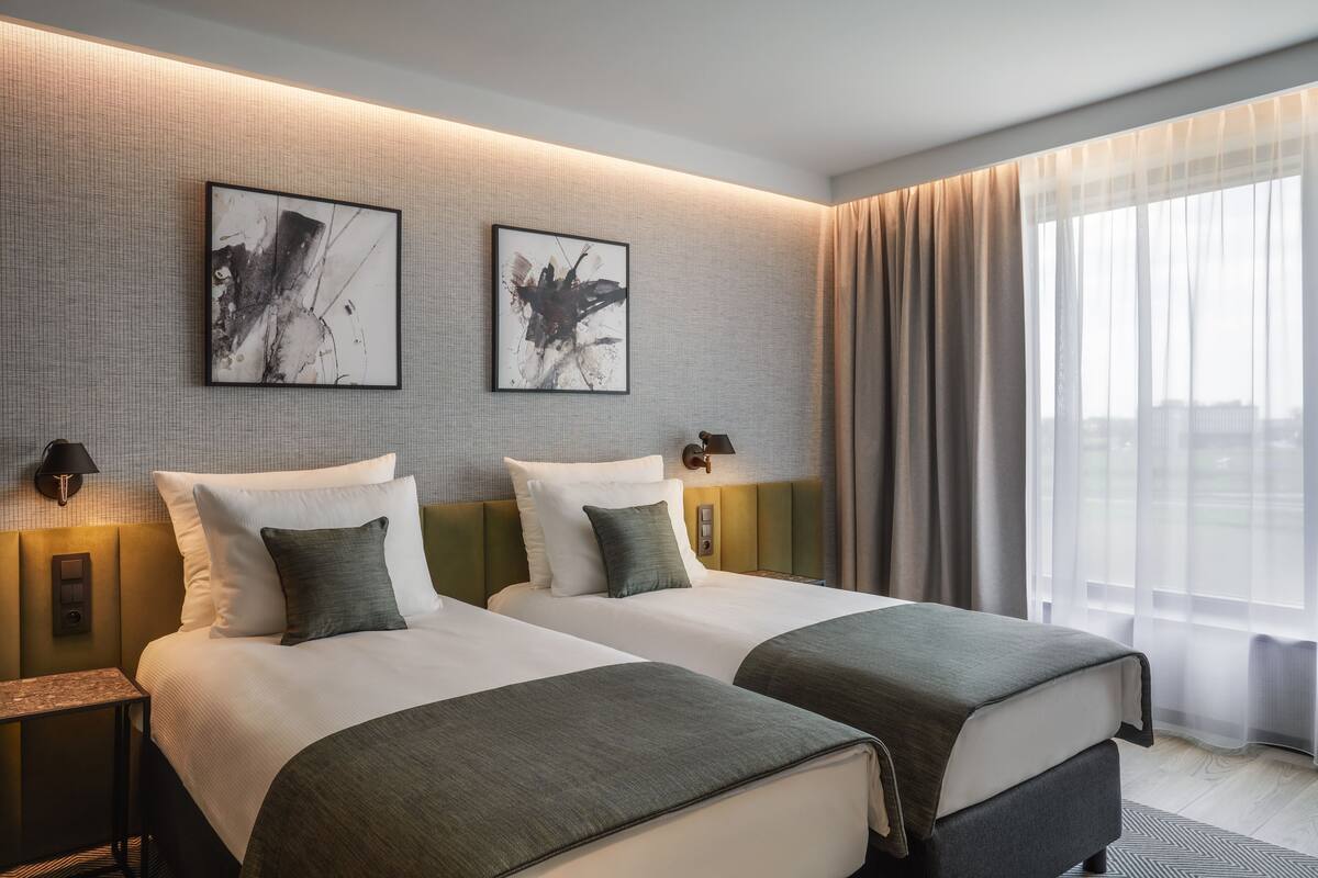 Quarto de hotel com duas camas, uma janela ampla com cortinas leves e arte abstrata nas paredes.