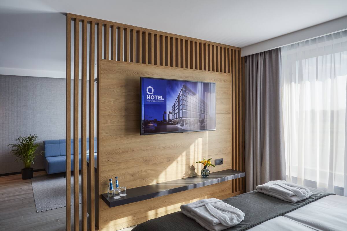Quarto de hotel moderno com uma televisão de tela plana grande fixada em um painel de madeira, cama com toalhas bem dobradas, mesinha de apoio com produtos de higiene pessoal e um vaso de planta.