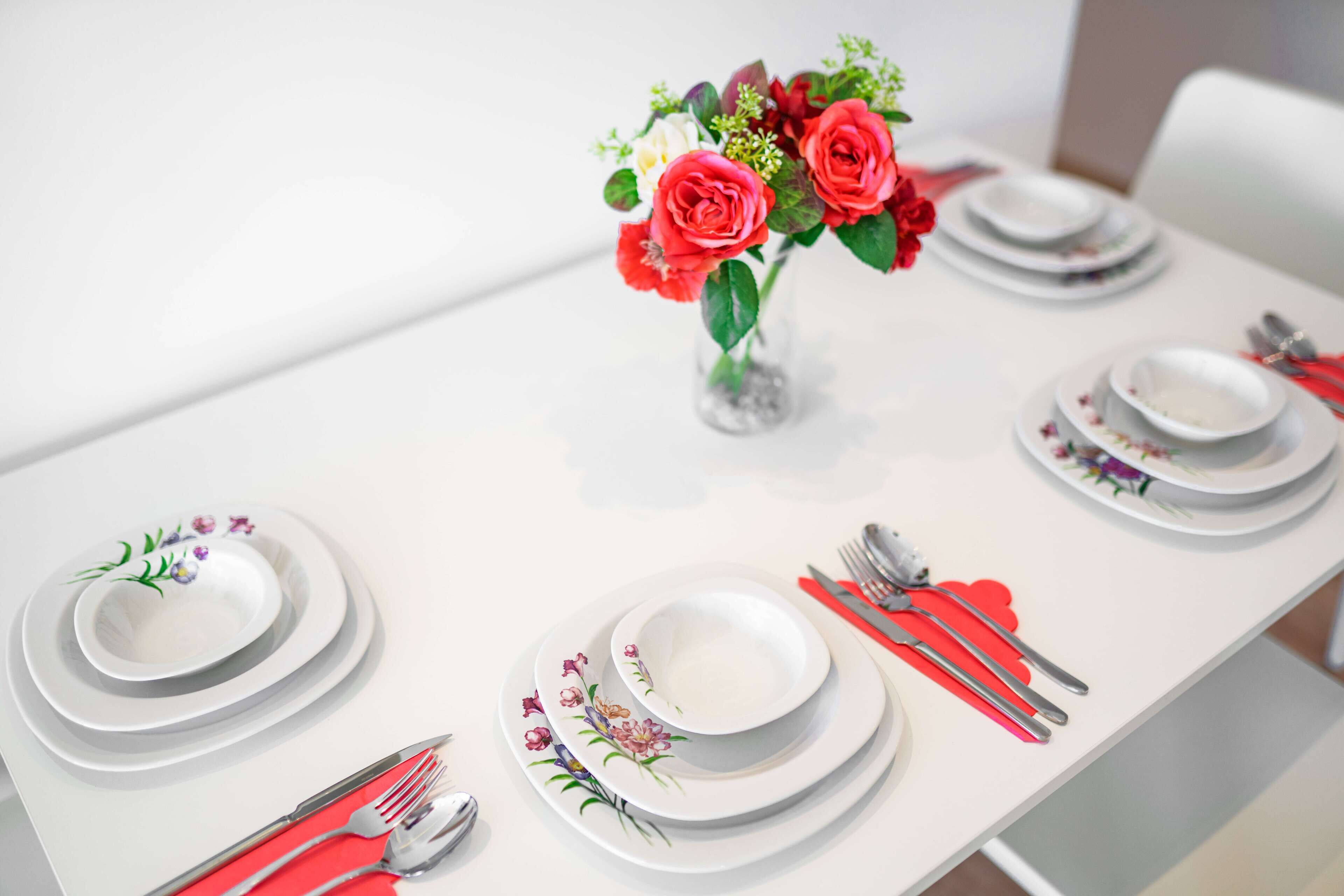 Mesa de jantar posta com pratos brancos, talheres vermelhos e um vaso de rosas vermelhas.