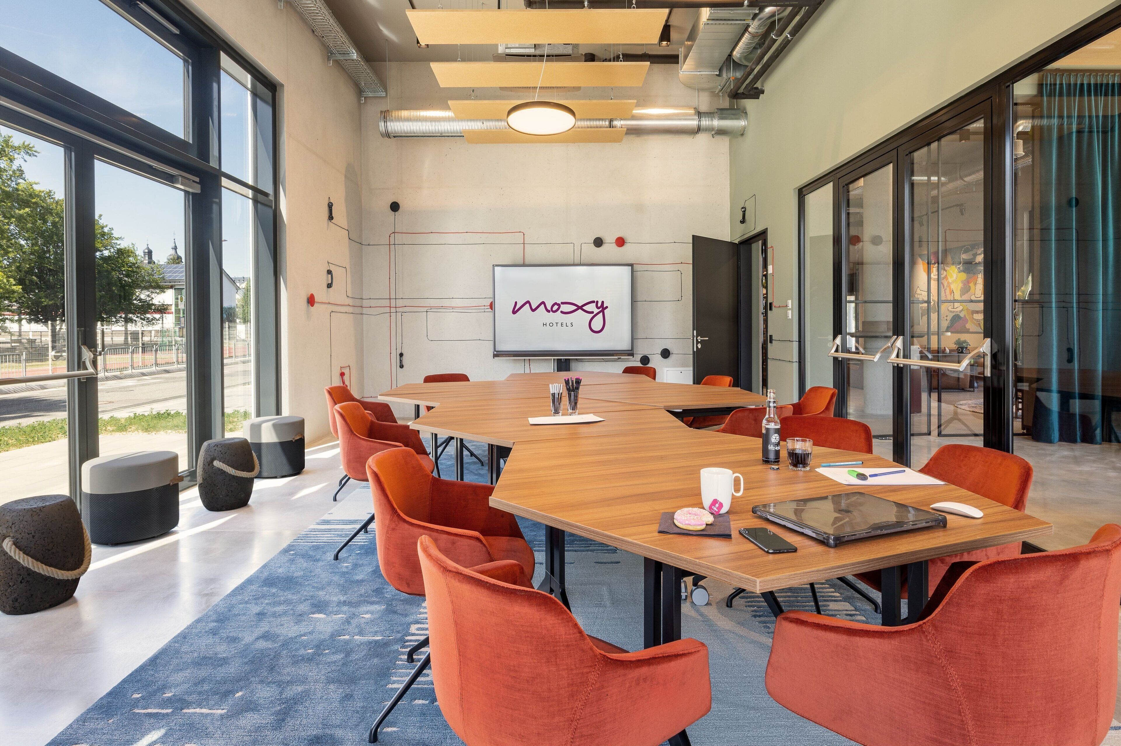 Uma sala de conferências com uma mesa grande, cadeiras laranja, um quadro branco com o logo "moosy hotels" e grandes janelas com vista para o exterior.