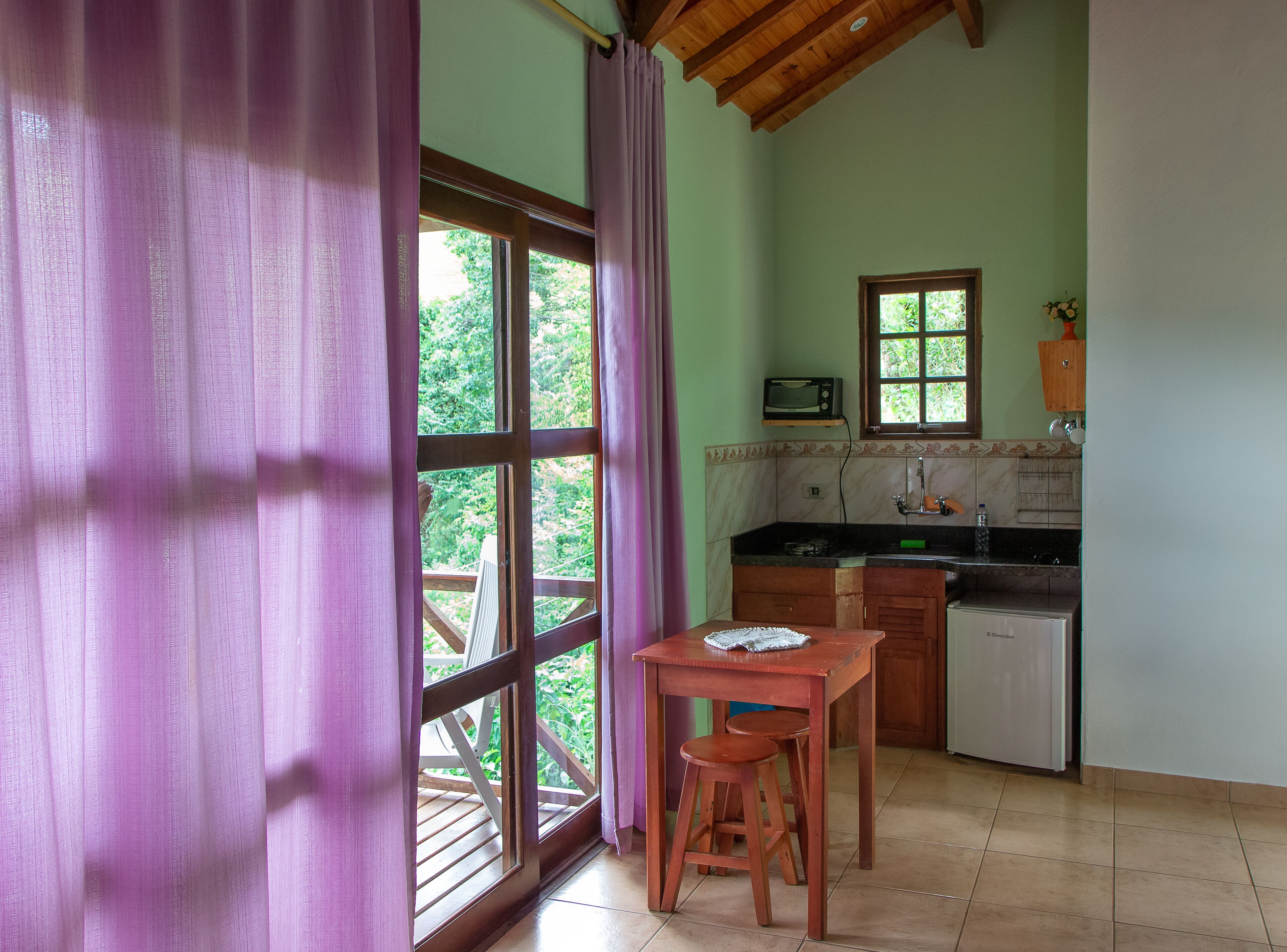 Cozinha com mesa de madeira e banquetas, janela com cortinas e vista para o verde lá fora.