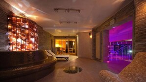 Uma área de spa com um elemento de pedras aquecidas, cadeiras de descanso e uma escadaria iluminada em roxo.