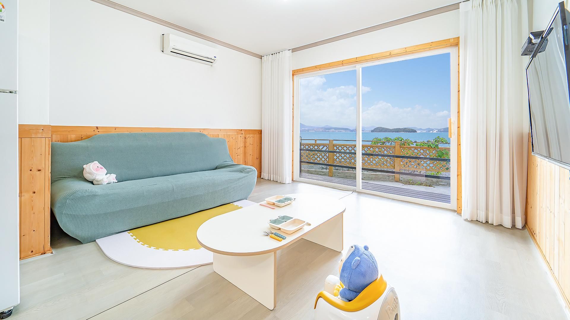 Quarto com sofá, mesa pequena, vista para o mar e janela com cortinas.
