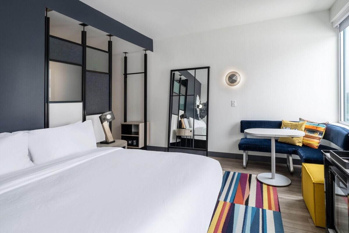Quarto de hotel moderno com uma cama grande, um sofá, uma mesinha e um espelho.