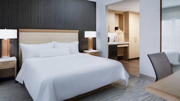 Quarto de hotel com uma cama grande, duas mesas de cabeceira com luminárias, uma escrivaninha com cadeira, e vista para o banheiro e a cozinha.