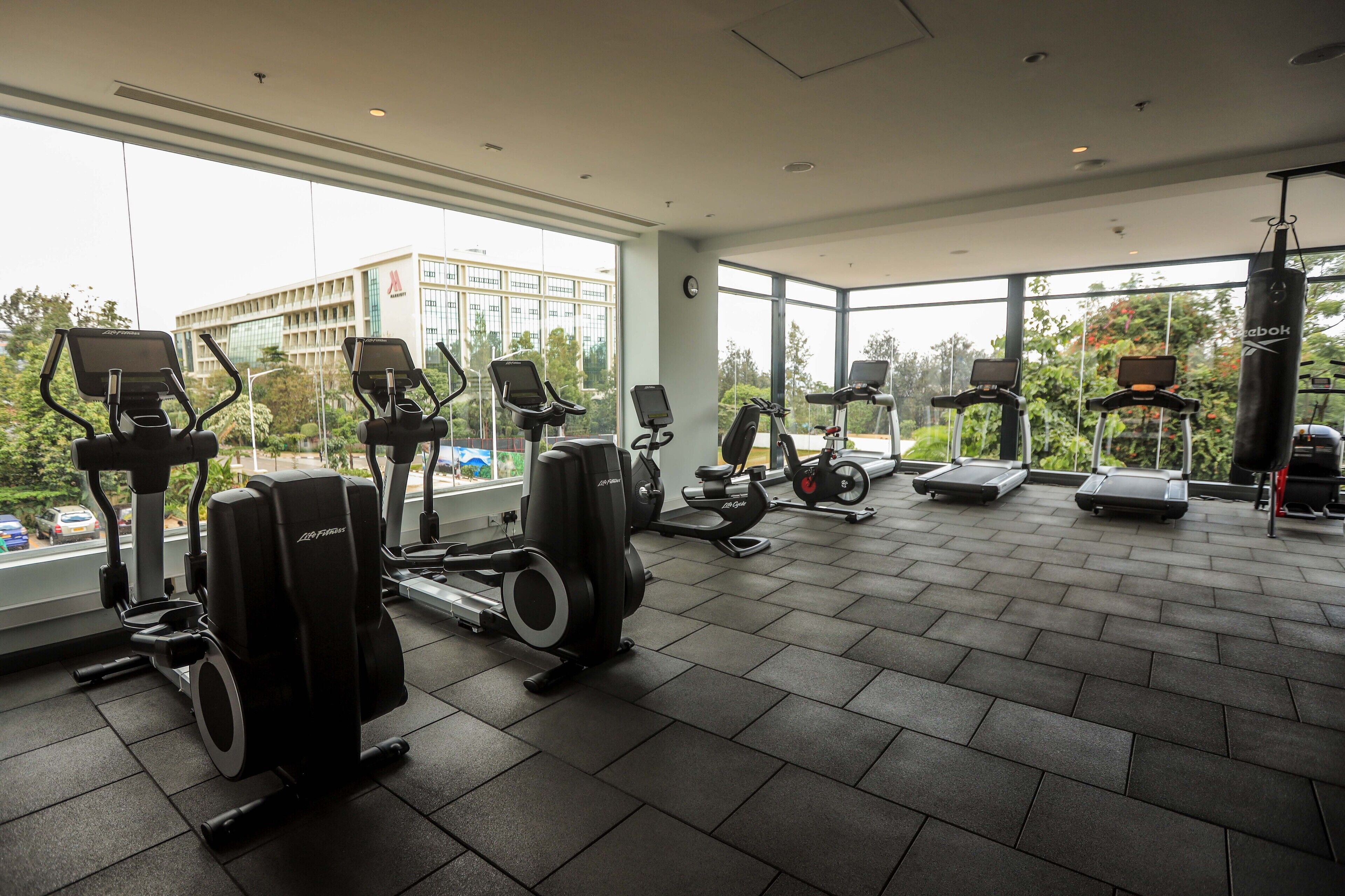 Uma academia com diversos equipamentos de exercício, janelas amplas e vista para um edifício.