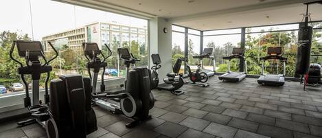 Uma academia com diversos equipamentos de exercício, janelas amplas e vista para um edifício.
